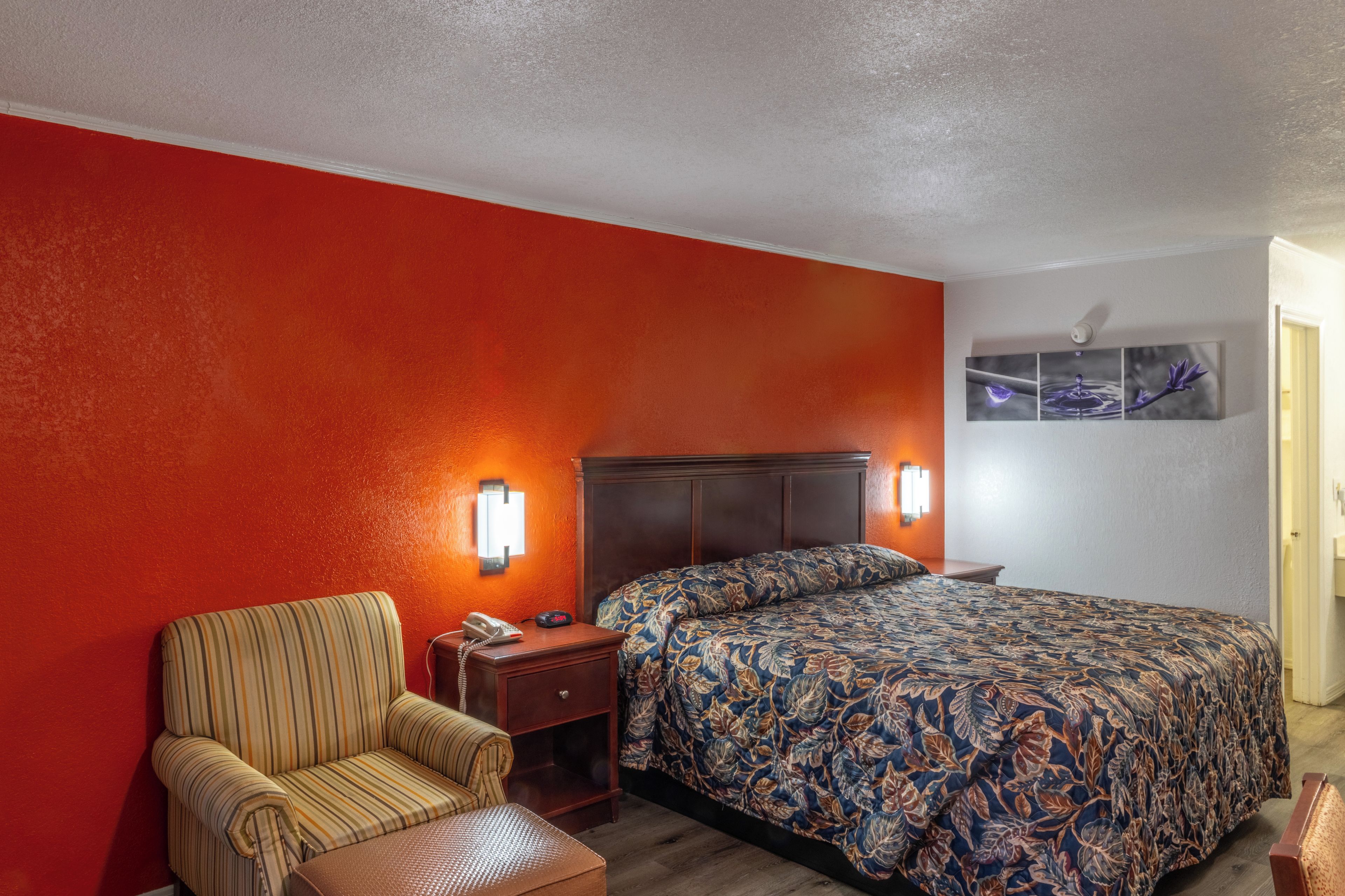 Quarto de hotel com parede vermelha, cama com colcha estampada, poltrona listrada, mesa de cabeceira com telefone e despertador, e luminária fixada na parede.
