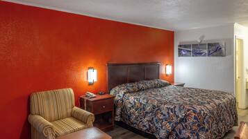 Quarto de hotel com parede vermelha, cama com colcha estampada, poltrona listrada, mesa de cabeceira com telefone e despertador, e luminária fixada na parede.