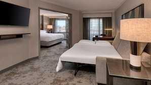 Quarto de hotel com uma cama grande, televisão na parede, escrivaninha com abajur e vista para a cidade pela janela.