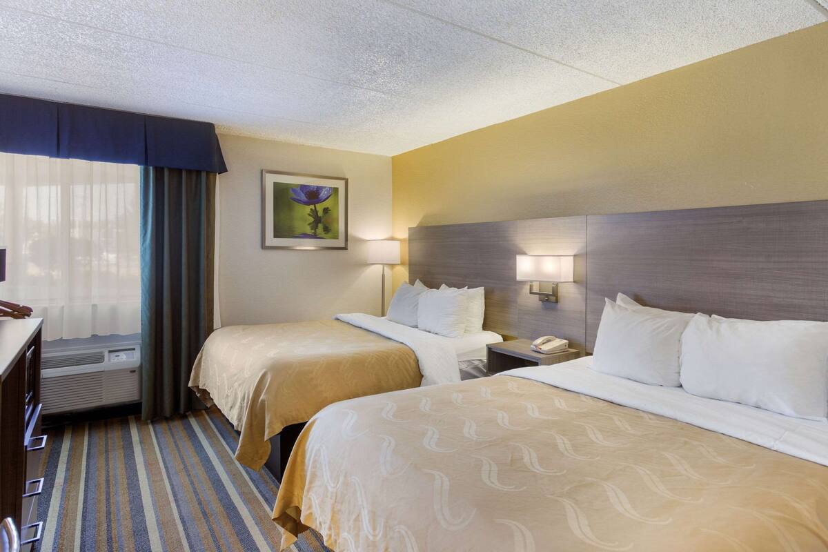 Quarto de hotel com duas camas, uma escrivaninha, uma cadeira, janela com cortinas, luminária fixada na parede e um quadro emoldurado.