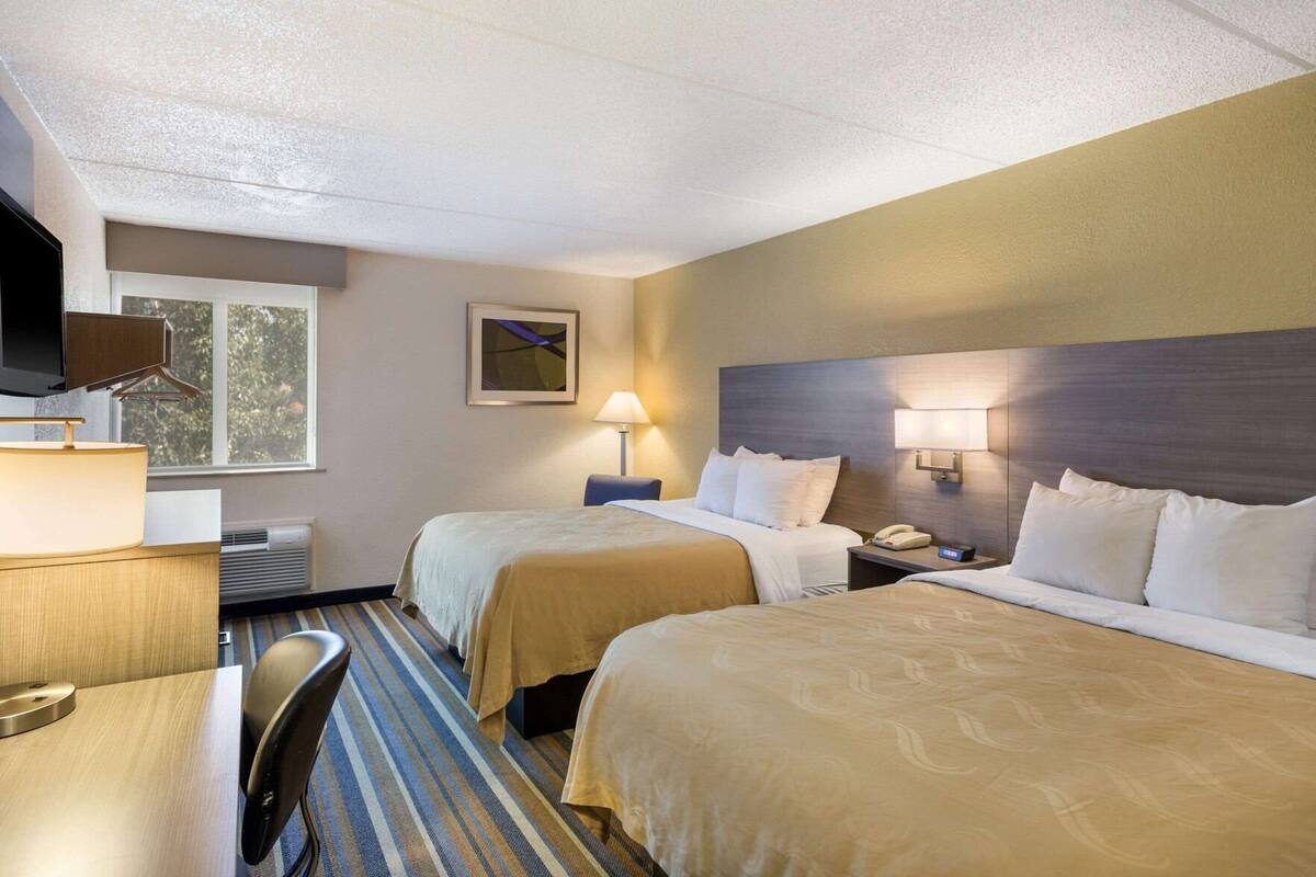 Quarto de hotel com duas camas, uma escrivaninha, uma cadeira, televisão, janela e um quadro na parede.