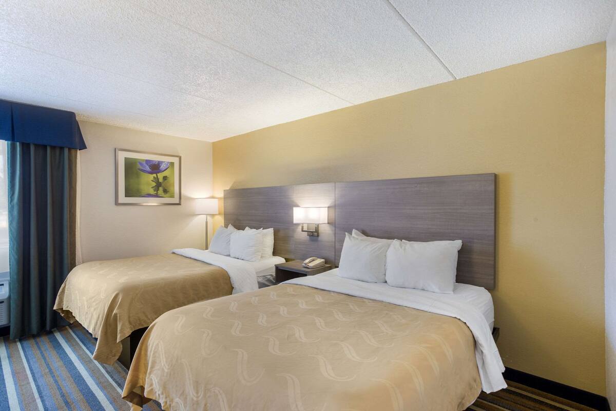 Quarto de hotel com duas camas, uma mesinha de cabeceira com abajur, um quadro na parede e uma janela com cortinas.