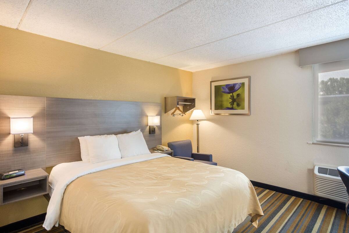 Quarto de hotel com uma cama grande, duas luminárias de cabeceira, uma cadeira, uma escrivaninha com telefone e uma janela.