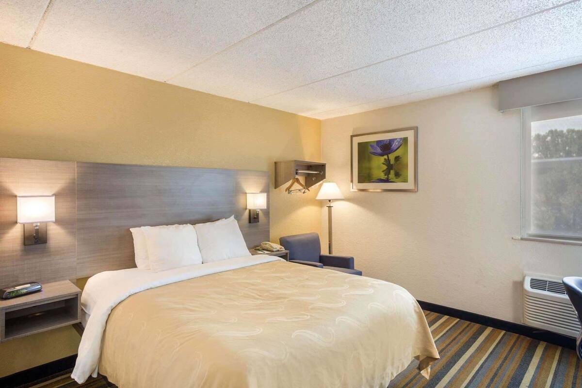 Quarto de hotel com uma cama grande, luminárias de cabeceira, escrivaninha com telefone, cadeira e janela.