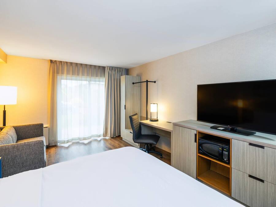 Quarto de hotel com cama, escrivaninha, cadeira, televisão e janela com cortinas.