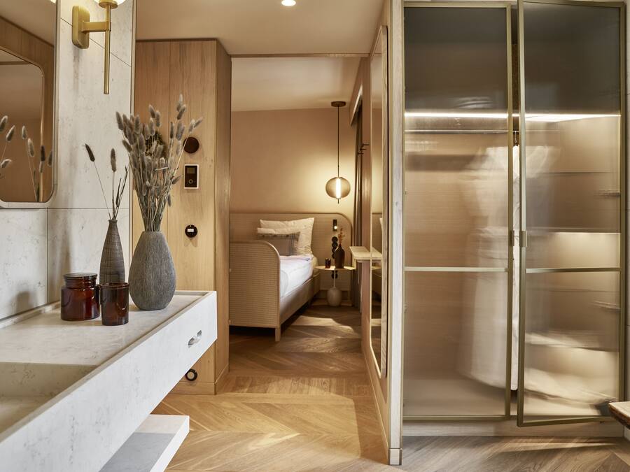 Quarto de hotel moderno com piso de madeira, um espelho grande, mesa de cabeceira com abajur e um guarda-roupa com portas de correr.