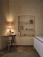 Quarto com uma escrivaninha de madeira, uma cadeira, mesa de cabeceira, abajur e estantes com livros e objetos decorativos.
