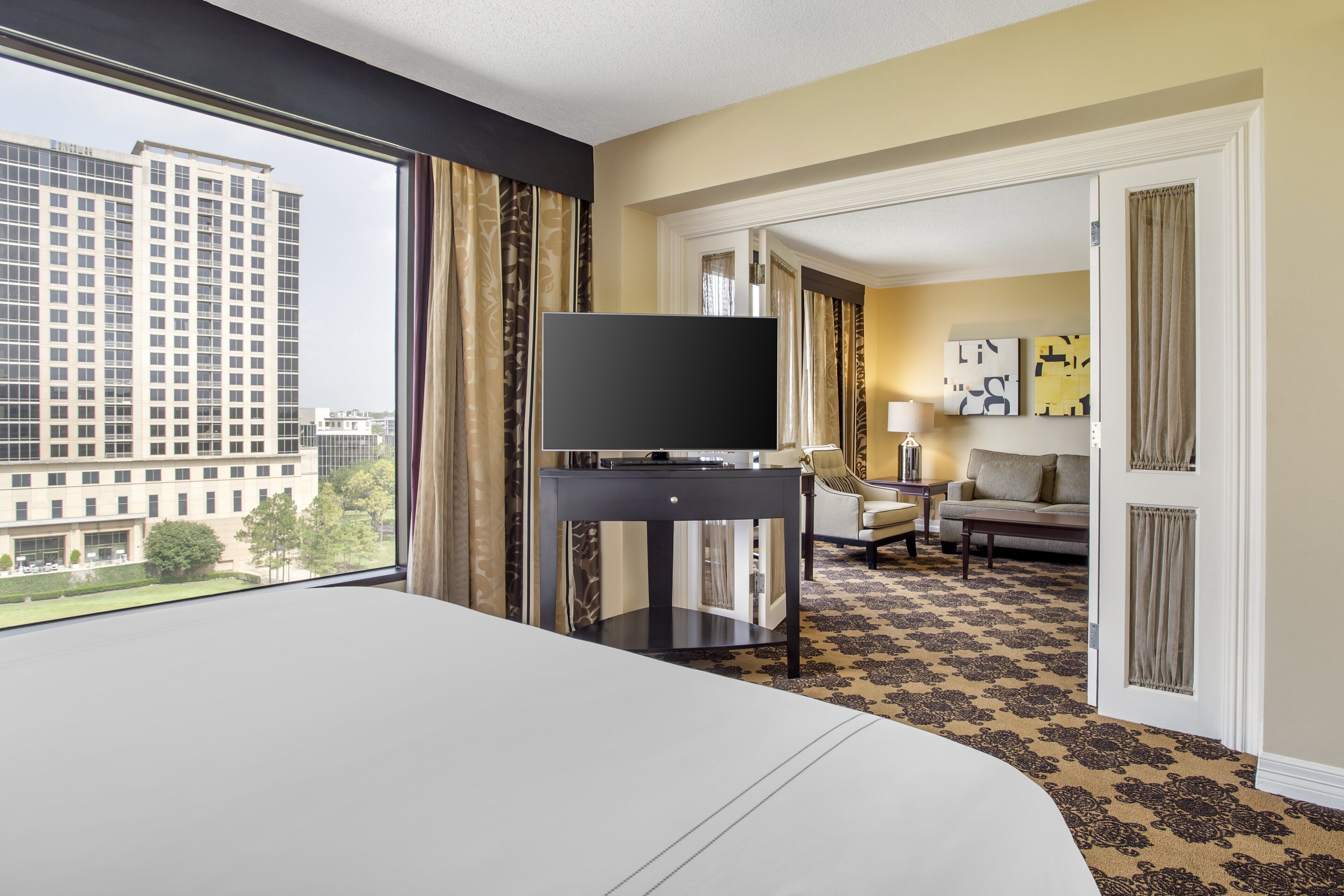 Quarto de hotel com uma cama grande, televisão em suporte, escrivaninha, sofá e vista para a paisagem urbana pela janela.