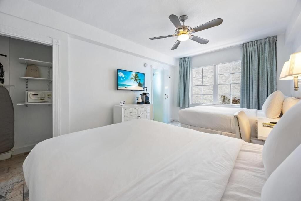 Quarto de hotel com cama, escrivaninha, cadeira, ventoinha no teto, televisão e janela com cortinas.
