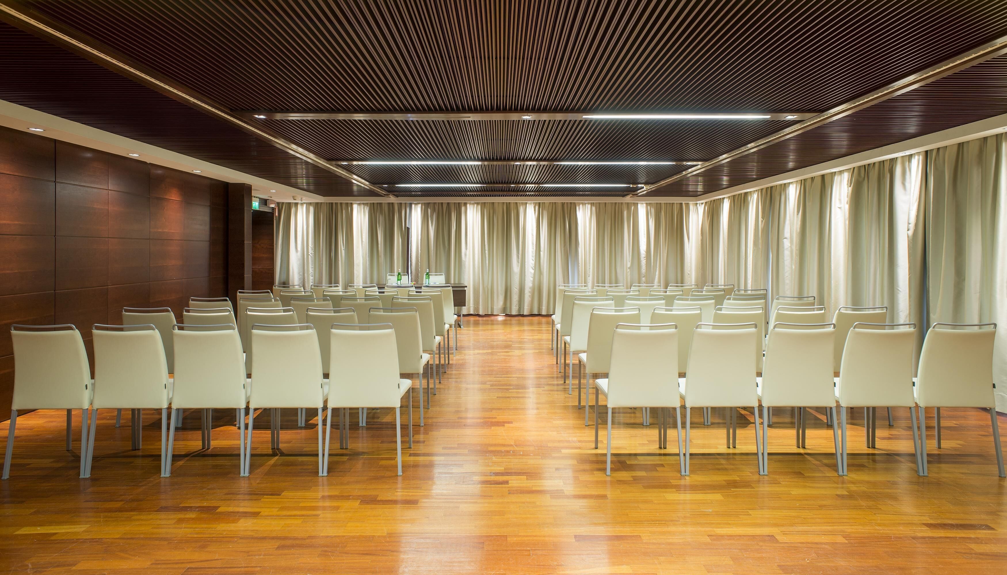 Uma sala de conferências com fileiras de cadeiras brancas, piso de madeira e teto com padrão linear.