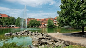 Um parque tranquilo com uma fonte, um lago e um edifício com telhados vermelhos ao fundo.
