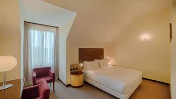 Quarto de hotel com cama, poltrona, abajur e mesa de cabeceira.
