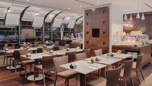 Um restaurante moderno com várias mesas e cadeiras, uma vitrine de vidro com itens alimentícios e uma grande janela com vista para a cidade.