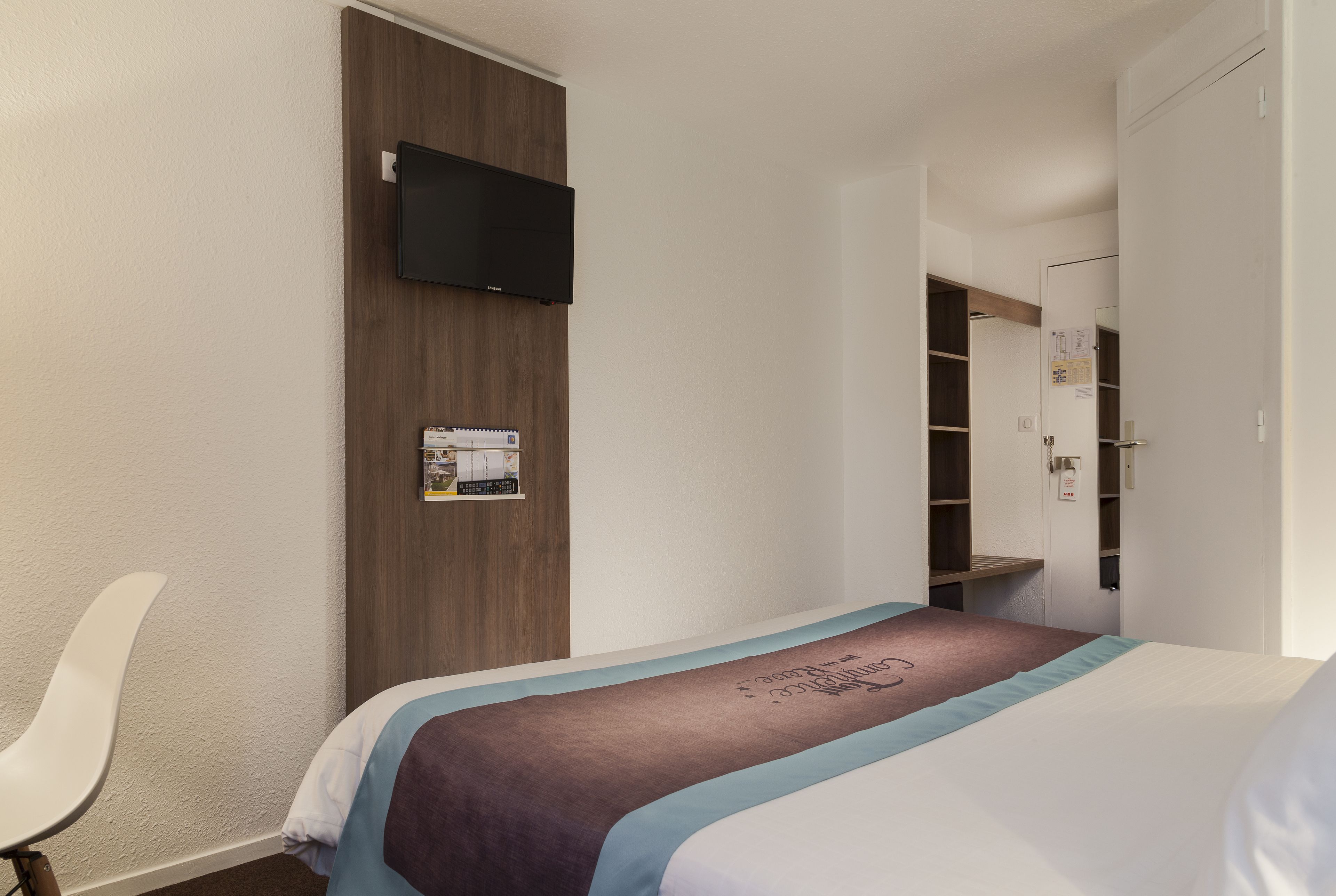 Quarto de hotel com cama, televisão fixada na parede, cadeira branca e guarda-roupa.