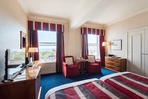 Quarto de hotel com uma cama grande, duas poltronas, uma escrivaninha com televisão e vista para o exterior.