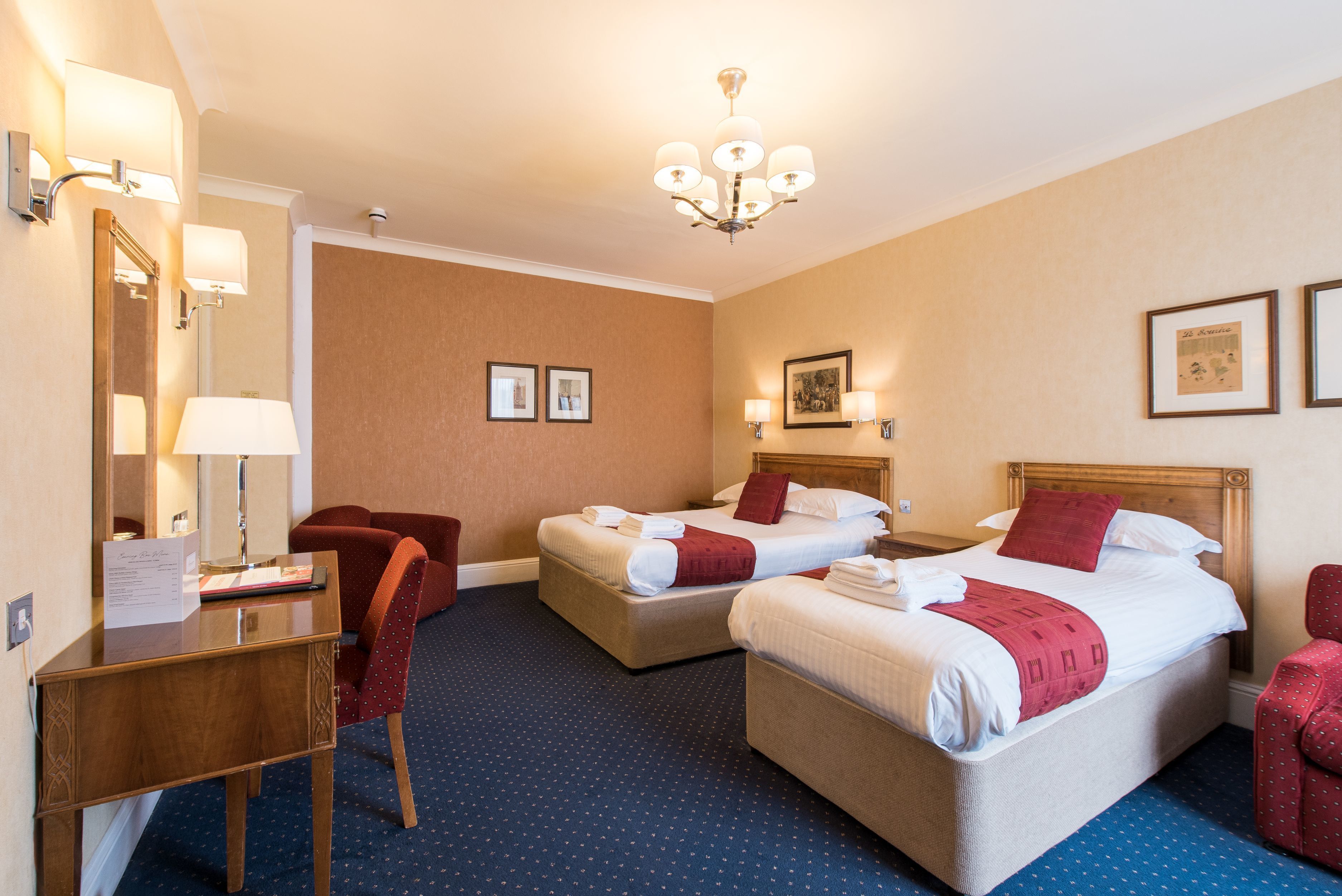 Quarto de hotel com duas camas, uma escrivaninha, uma cadeira e um candelabro.