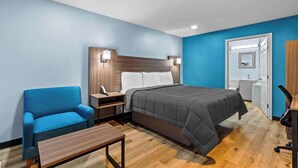 Quarto de hotel com uma cama, poltrona azul, mesa de cabeceira de madeira e uma escrivaninha com cadeira.