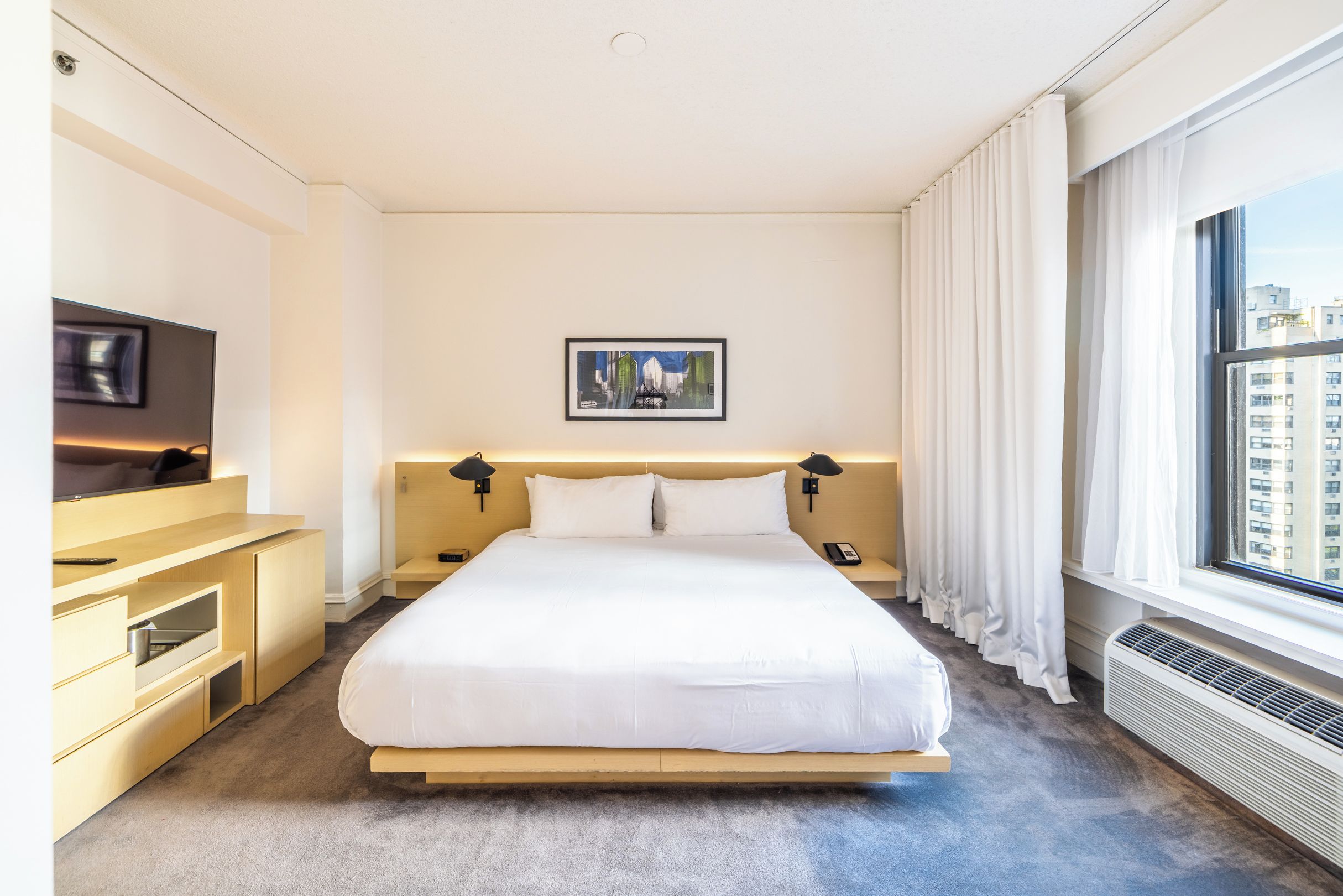 Quarto de hotel com cama, televisão, um quadro, janela com cortinas e vista para prédios.
