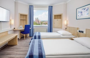 Quarto de hotel com duas camas, uma escrivaninha com cadeira, televisão e janela com vista para uma igreja.