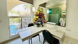 Quarto de hotel moderno com área de refeições, uma janela grande com vista para uma piscina e palmeiras, um espelho e um vaso de flores.
