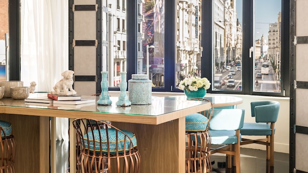 Uma área de refeições moderna com mesa de vidro, cadeiras de vime e vista para a cidade através de grandes janelas.
