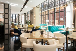 Um hall de hotel moderno com uma mesa de centro de vidro, assentos em branco e azul, e grandes janelas com vista para a rua.