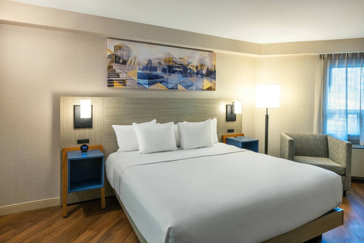 Quarto de hotel com uma cama grande, duas mesas de cabeceira, uma cadeira, um quadro na parede e uma janela com cortinas.