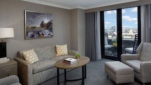 Quarto de hotel moderno com sofá cinzento, poltrona a combinar, mesa redonda pequena com um livro e uma planta, e uma janela grande com vista para a cidade.