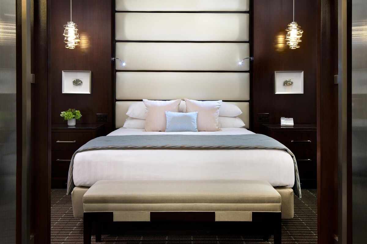 Quarto de hotel com uma cama grande, mesas de cabeceira, um banco e iluminação na parede.