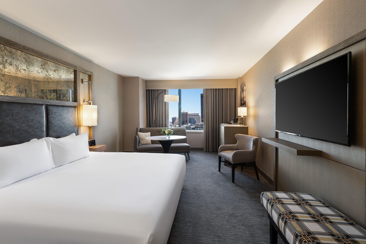 Quarto de hotel com uma cama grande, área de estar com mesa, televisão e vista da cidade.