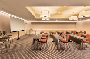 Sala de conferências com tela de projetor, mesas, cadeiras e iluminação pendente.
