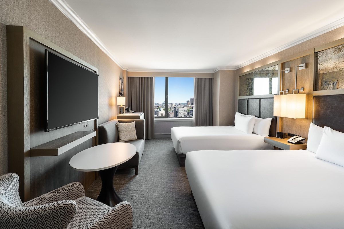Quarto de hotel com duas camas, televisão, escrivaninha e vista para a cidade.
