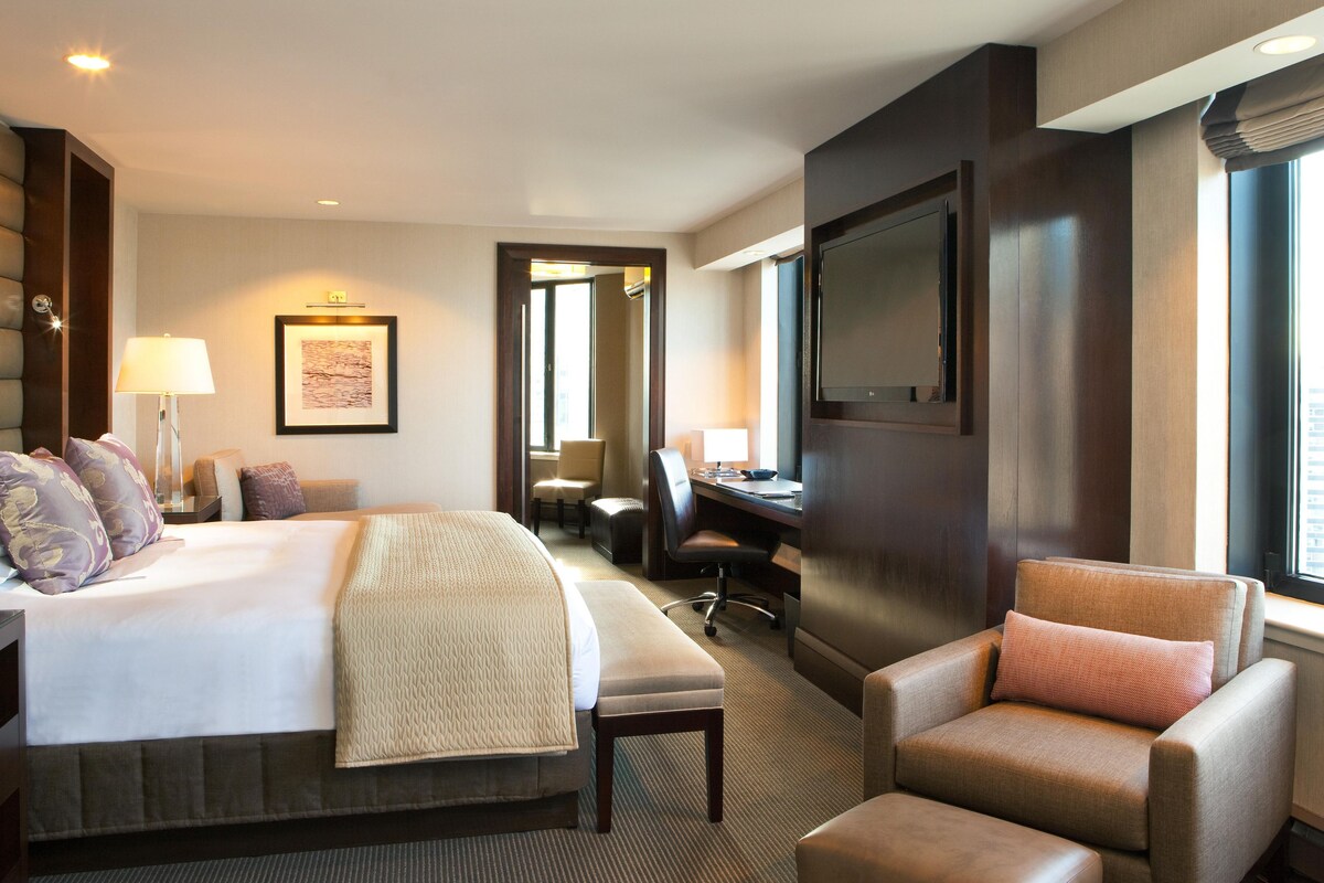 Quarto de hotel com cama, escrivaninha, cadeira e televisão.