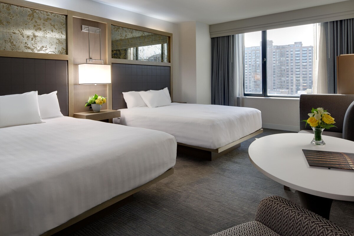 Quarto de hotel com duas camas, uma escrivaninha, uma cadeira e vista para a cidade.