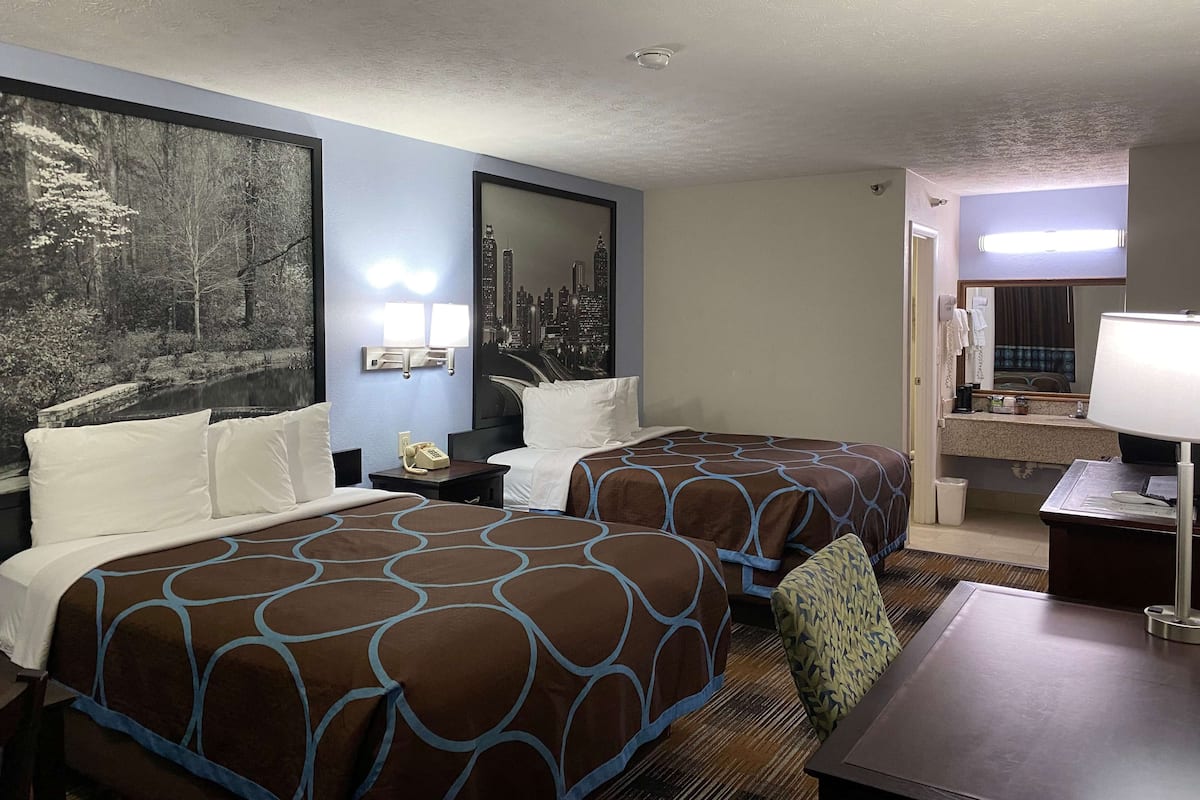 Quarto de hotel com duas camas, uma escrivaninha, uma cadeira, um espelho e um banheiro visível através de uma porta aberta.