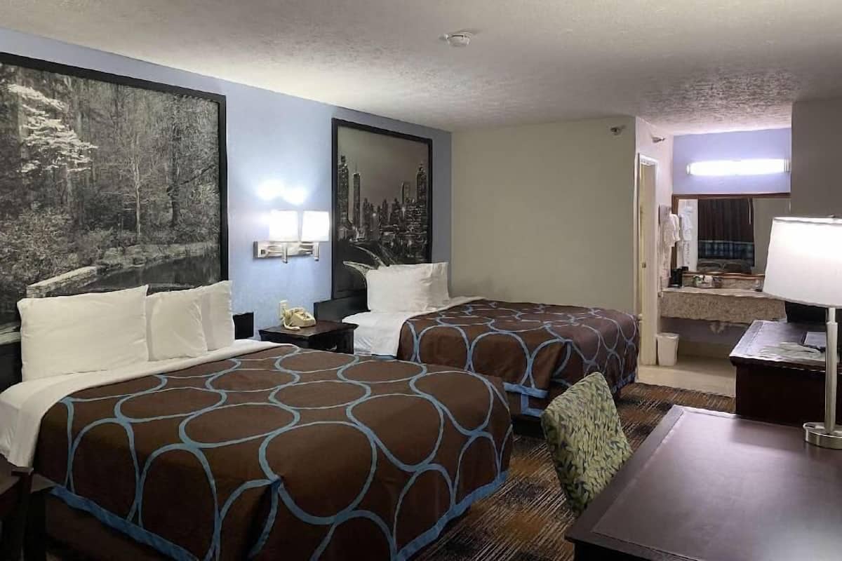 Quarto de hotel com duas camas, uma escrivaninha, uma cadeira, um espelho e banheiro com pia e suporte para toalhas.