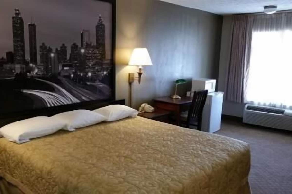 Quarto de hotel com cama, duas luminárias de cabeceira, escrivaninha com cadeira, computador e janela com cortinas.