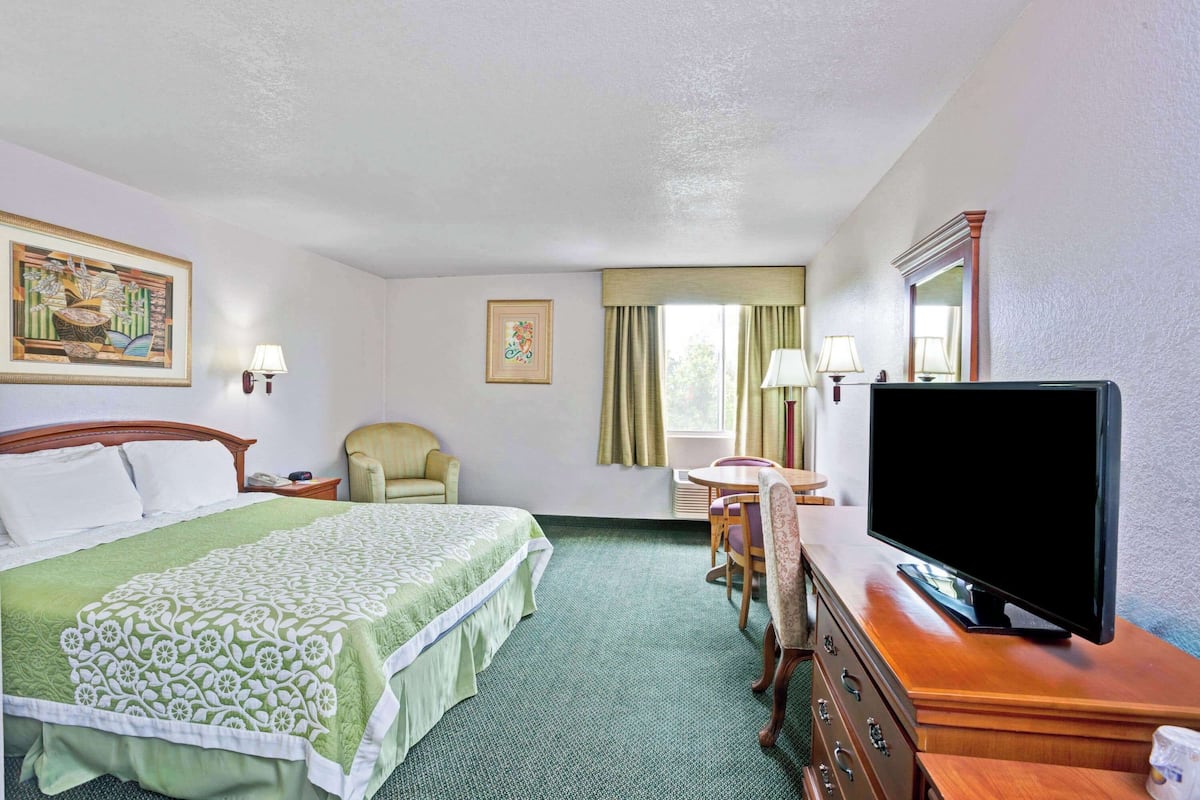 Quarto de hotel com cama, televisão, escrivaninha com cadeira, abajur e janela com cortinas.