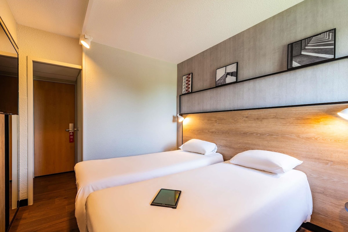 Quarto de hotel com duas camas, cabeceira de madeira, mesa de cabeceira, um tablet na cama e uma porta com uma placa vermelha.