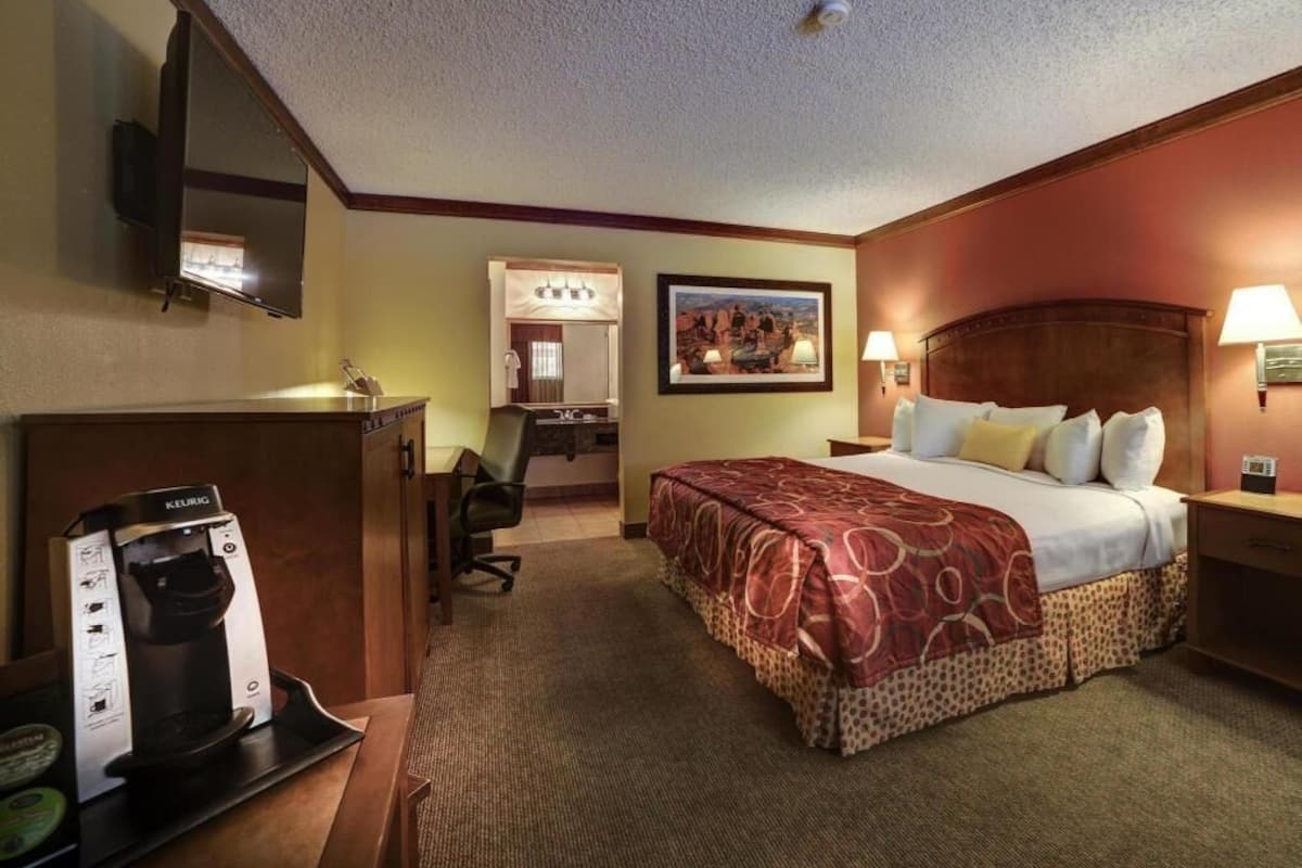 Quarto de hotel com cama, escrivaninha, cadeira, cafeteira e um quadro na parede.