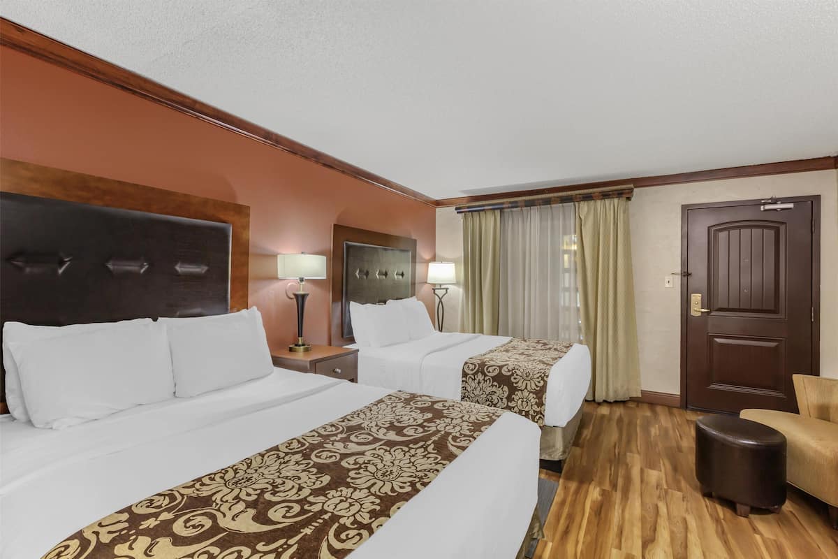 Quarto de hotel com duas camas, piso de madeira, porta marrom e um banquinho redondo pequeno.