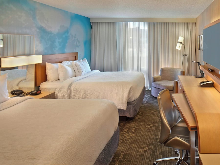 Quarto de hotel com duas camas, uma escrivaninha, uma cadeira, uma televisão e um abajur.