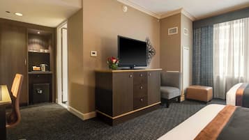 Quarto de hotel com uma cama grande, uma TV de tela plana sobre um móvel de madeira, uma escrivaninha com cadeira, um cofre e uma pequena área de estar.