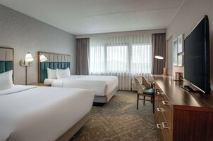 Quarto de hotel com duas camas, uma secretária com televisão e uma janela grande com cortinas.