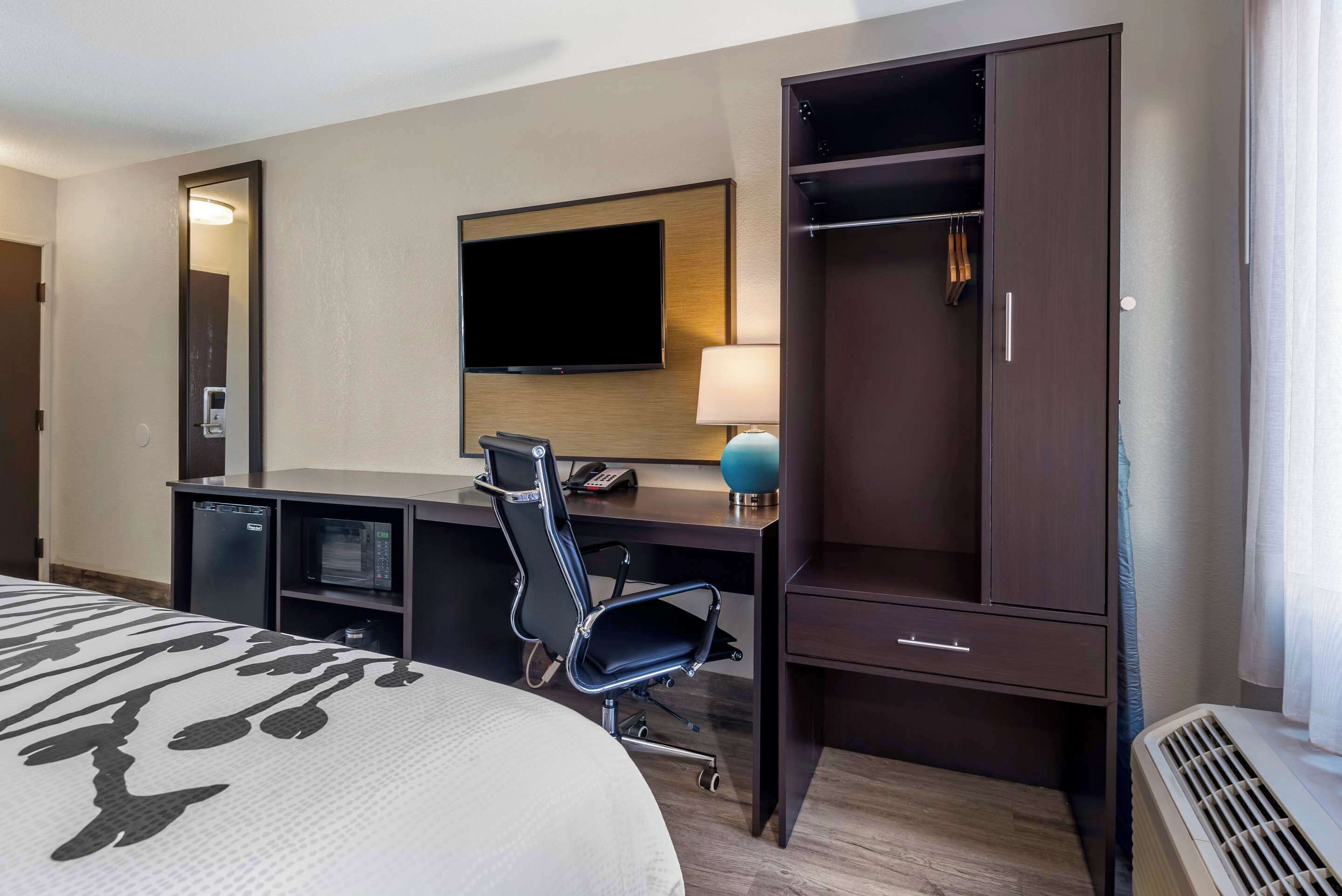 Quarto de hotel com cama, escrivaninha com computador, cadeira, guarda-roupa, televisão e abajur.