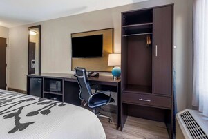 Quarto de hotel com cama, escrivaninha com computador, cadeira, guarda-roupa, televisão e abajur.