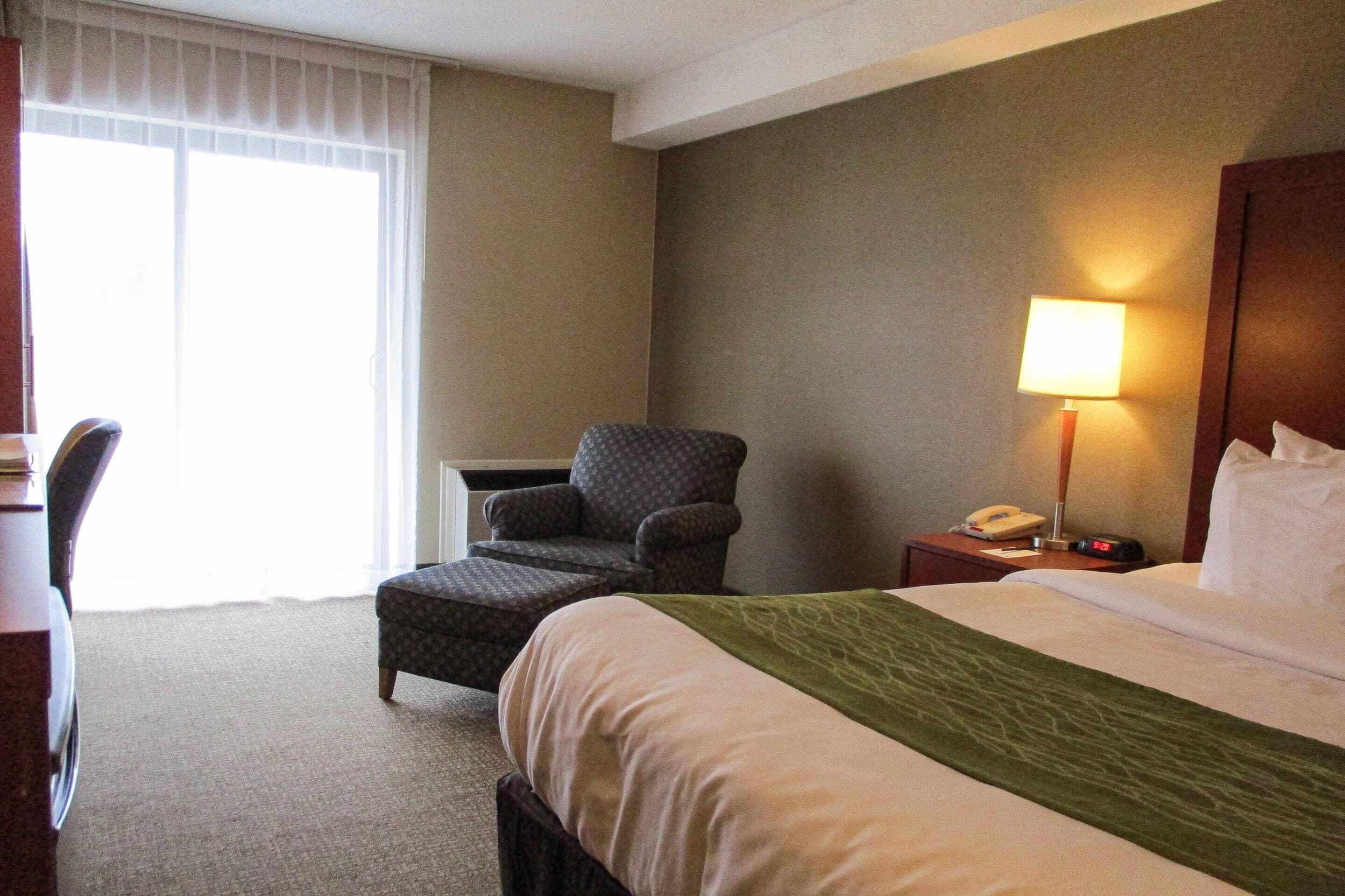Quarto de hotel com cama, cadeira, escrivaninha, abajur e janela com cortinas.