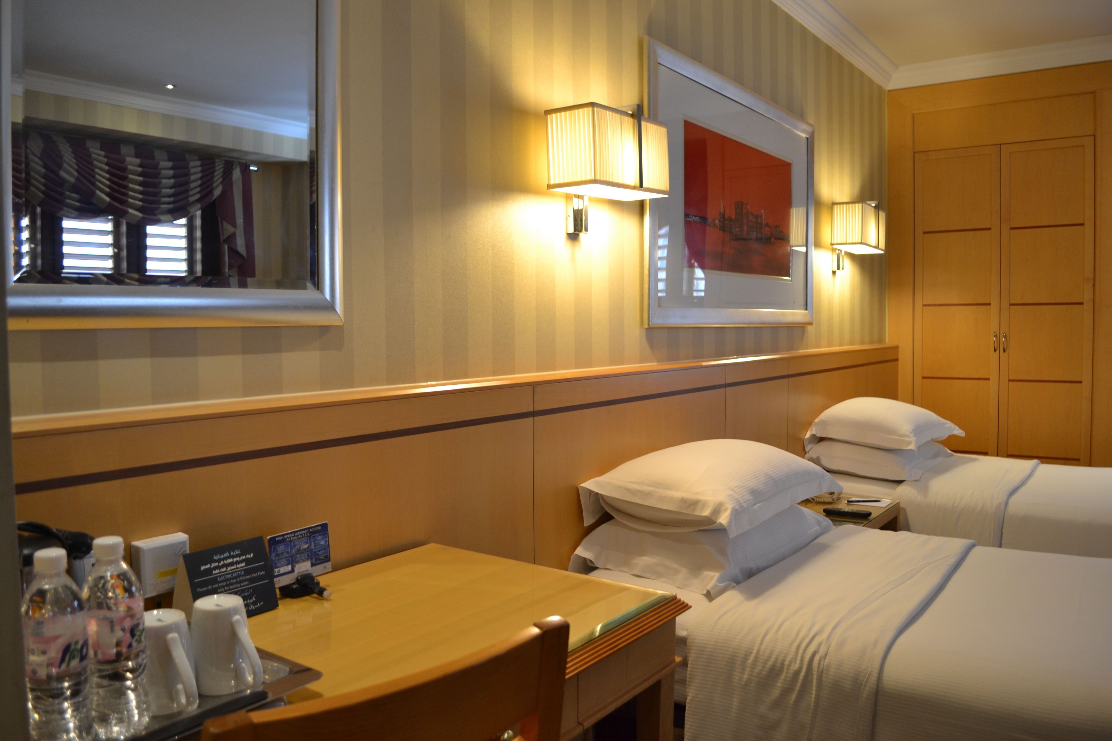 Quarto de hotel com duas camas, uma escrivaninha e uma janela com cortinas. Há luminárias fixadas na parede e um quadro emoldurado sobre as camas.