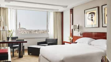 Quarto de hotel com uma cama grande, uma escrivaninha com cadeira, uma mesinha de cabeceira com abajur e vista para a paisagem urbana através de uma janela ampla.