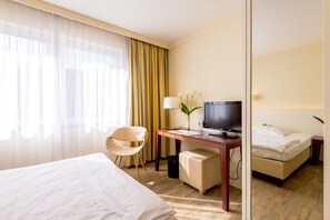 Quarto de hotel com uma cama, uma mesa de trabalho com televisão, uma cadeira e uma pequena mesa com luminária e flores.